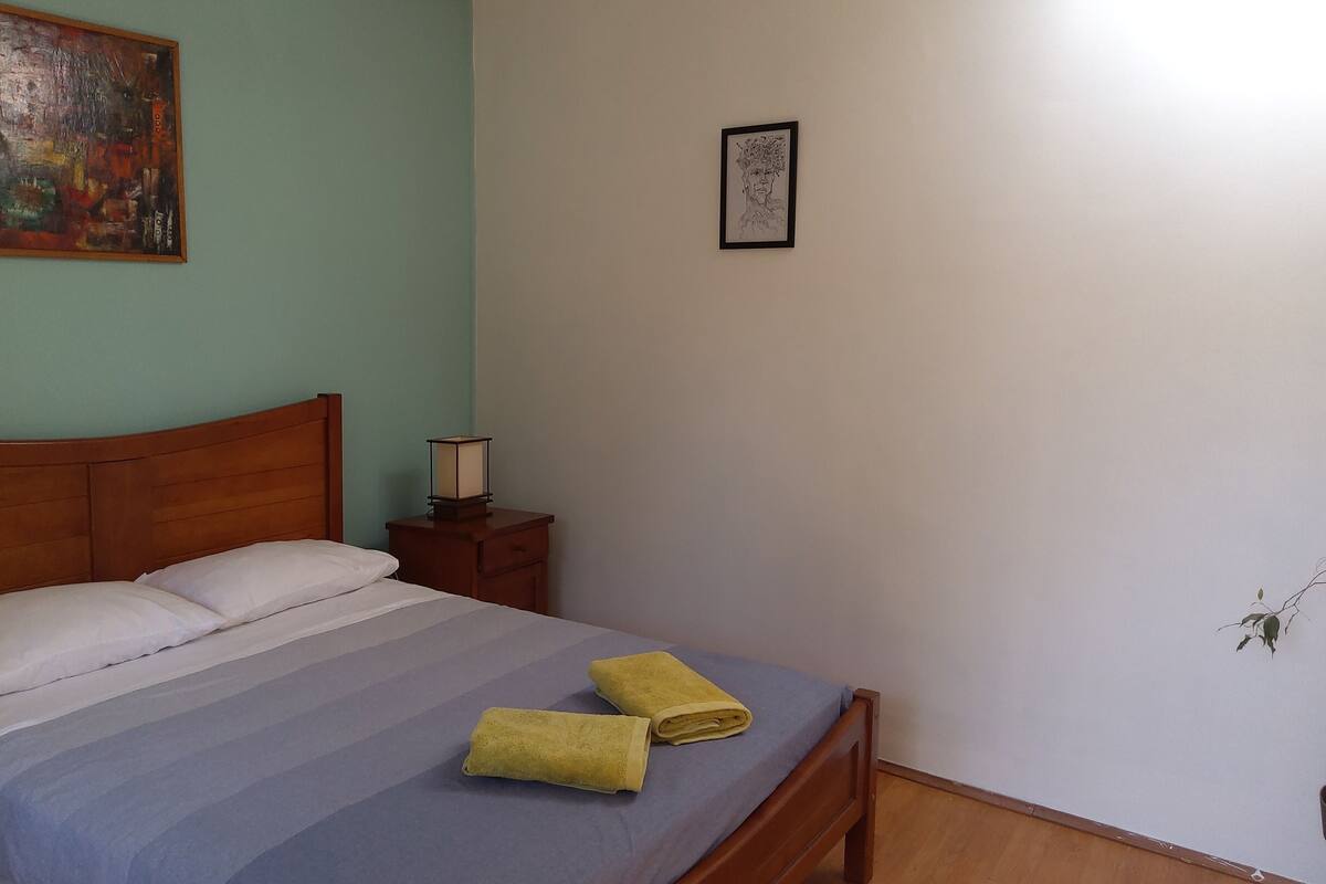 Cama bem arrumada com colcha listrada em azul e branco, duas toalhas amarelas, cabeceira de madeira, mesa de cabeceira com abajur e quadro pendurado na parede.