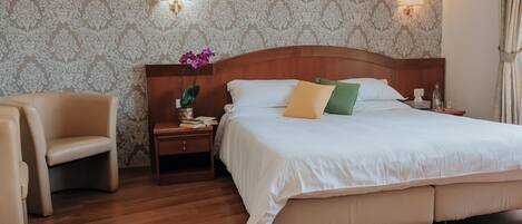 Quarto de hotel com cama de madeira, mesa de cabeceira com abajur, uma cadeira e papel de parede estampado.