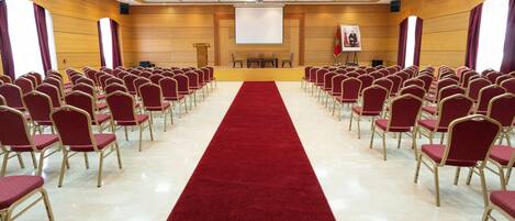 Uma sala de conferências com fileiras de cadeiras vermelhas, um tapete vermelho e uma tela de projetor na frente.