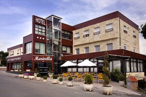 Um hotel com fachada moderna, restaurante à vista, área externa com mesas e guarda-sóis, e plantas em vasos.