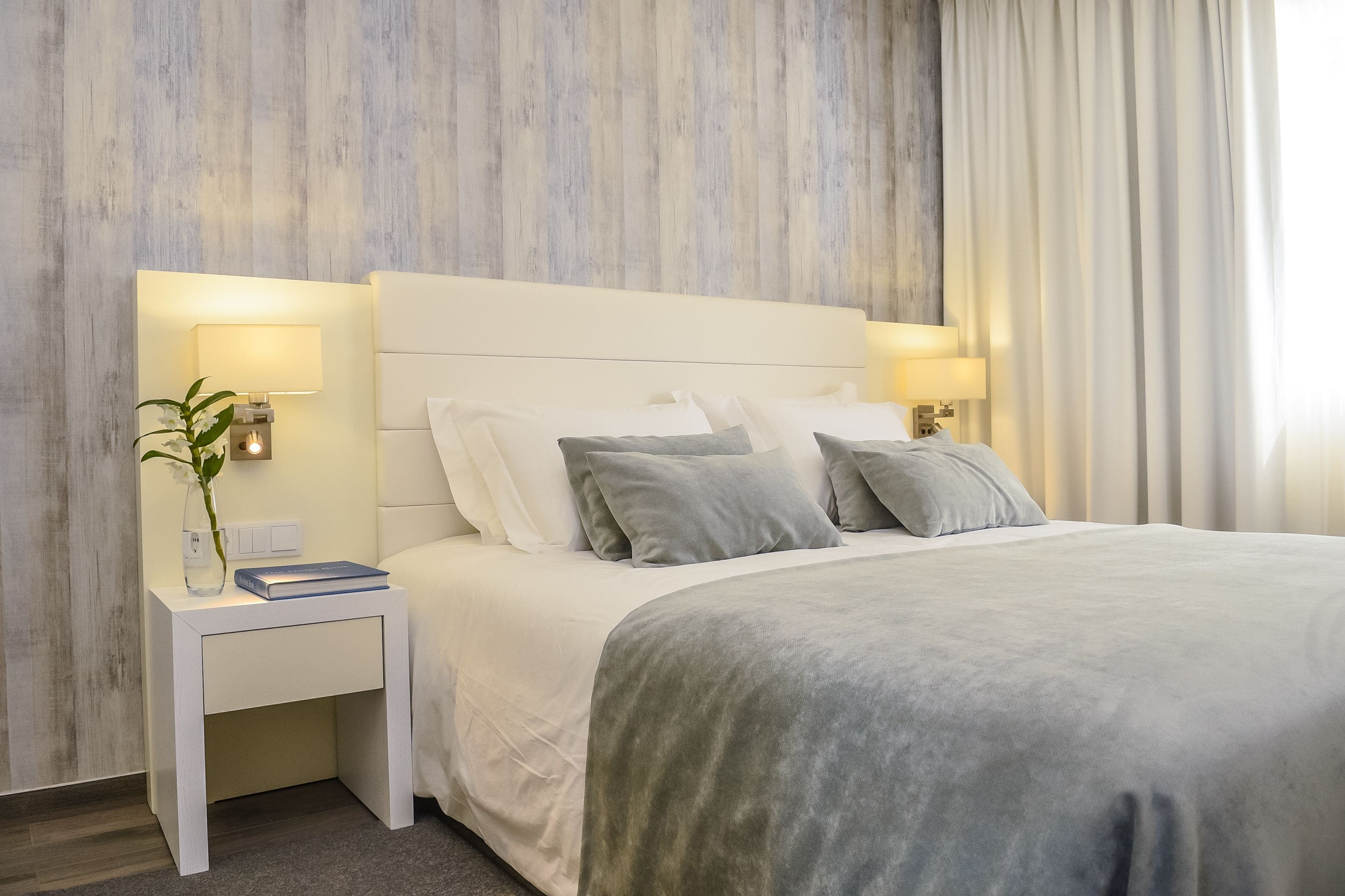 Cama bem arrumada com manta cinza e almofadas, duas luminárias de cabeceira, uma mesinha com um vaso e um livro, e uma parede de madeira com cortina.