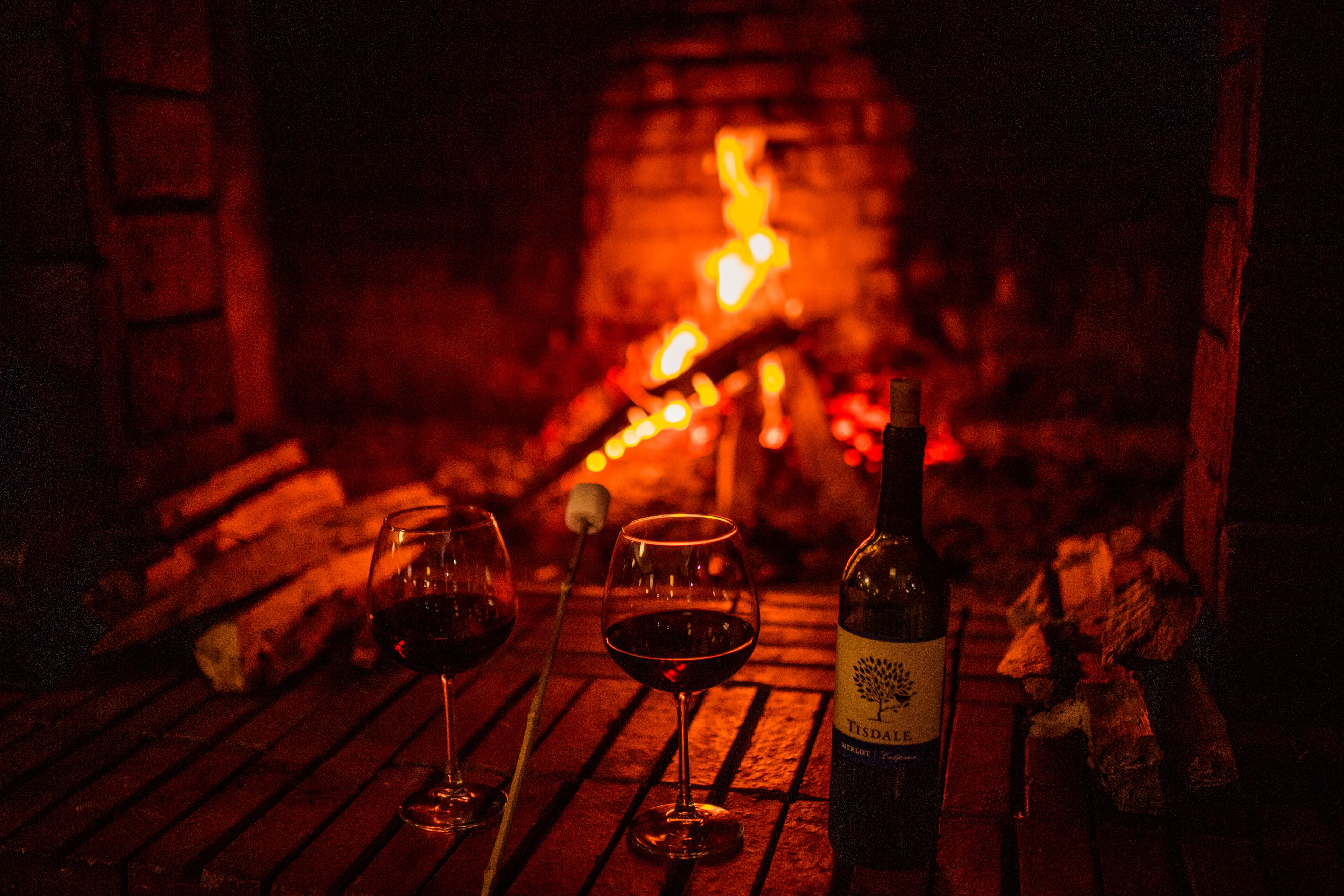 Mesa de madeira com duas taças de vinho e uma garrafa, com um cenário de lareira de tijolos e fogo.