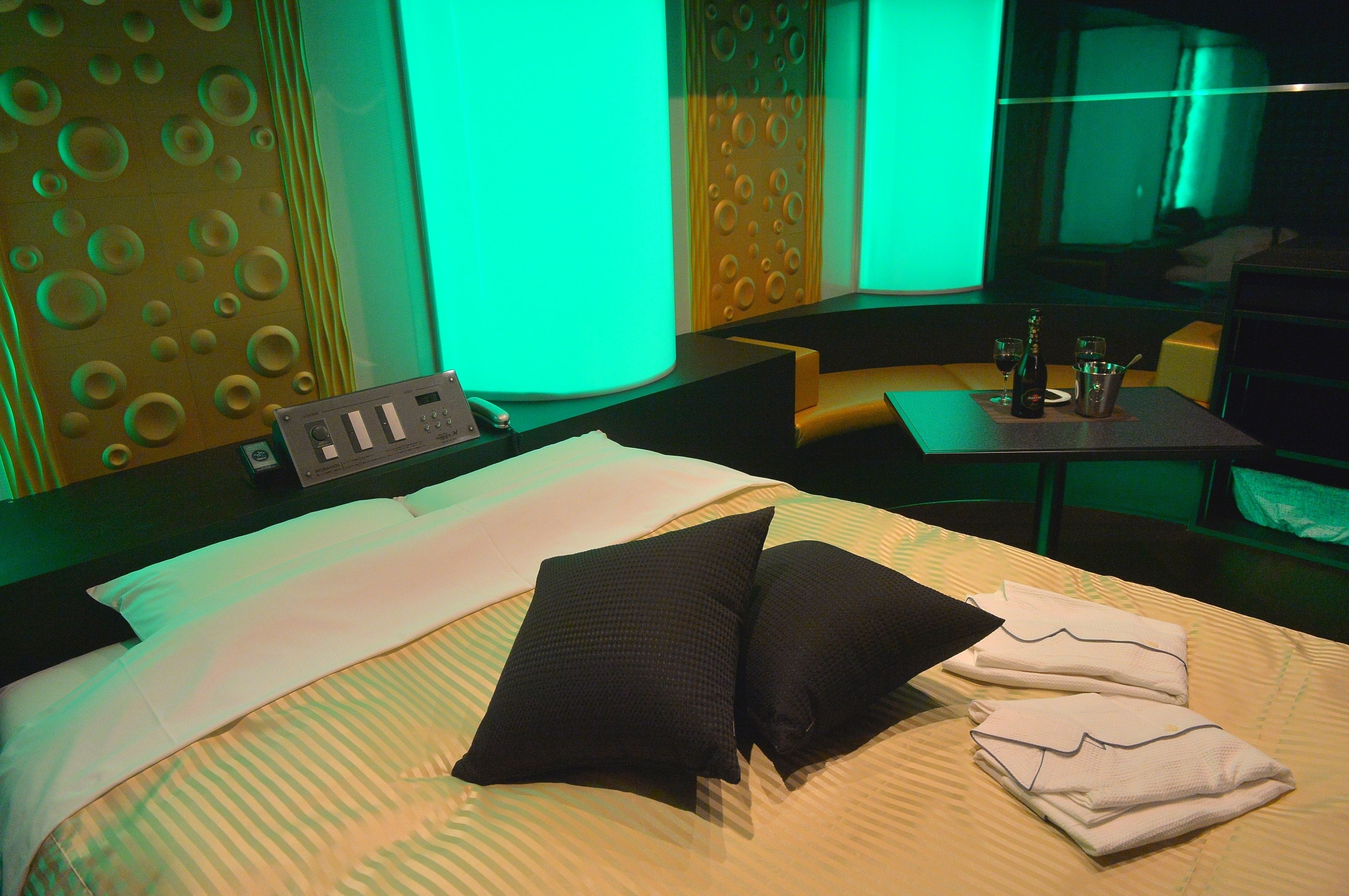 Quarto de hotel com cama, almofadas, mesa de cabeceira com garrafa e copos, e vista para o exterior através de uma janela.