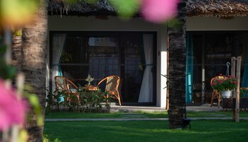 Uma área externa com assentos, incluindo duas cadeiras de vime e uma mesa, cercada por vegetação e uma estrutura de telhado de palha.