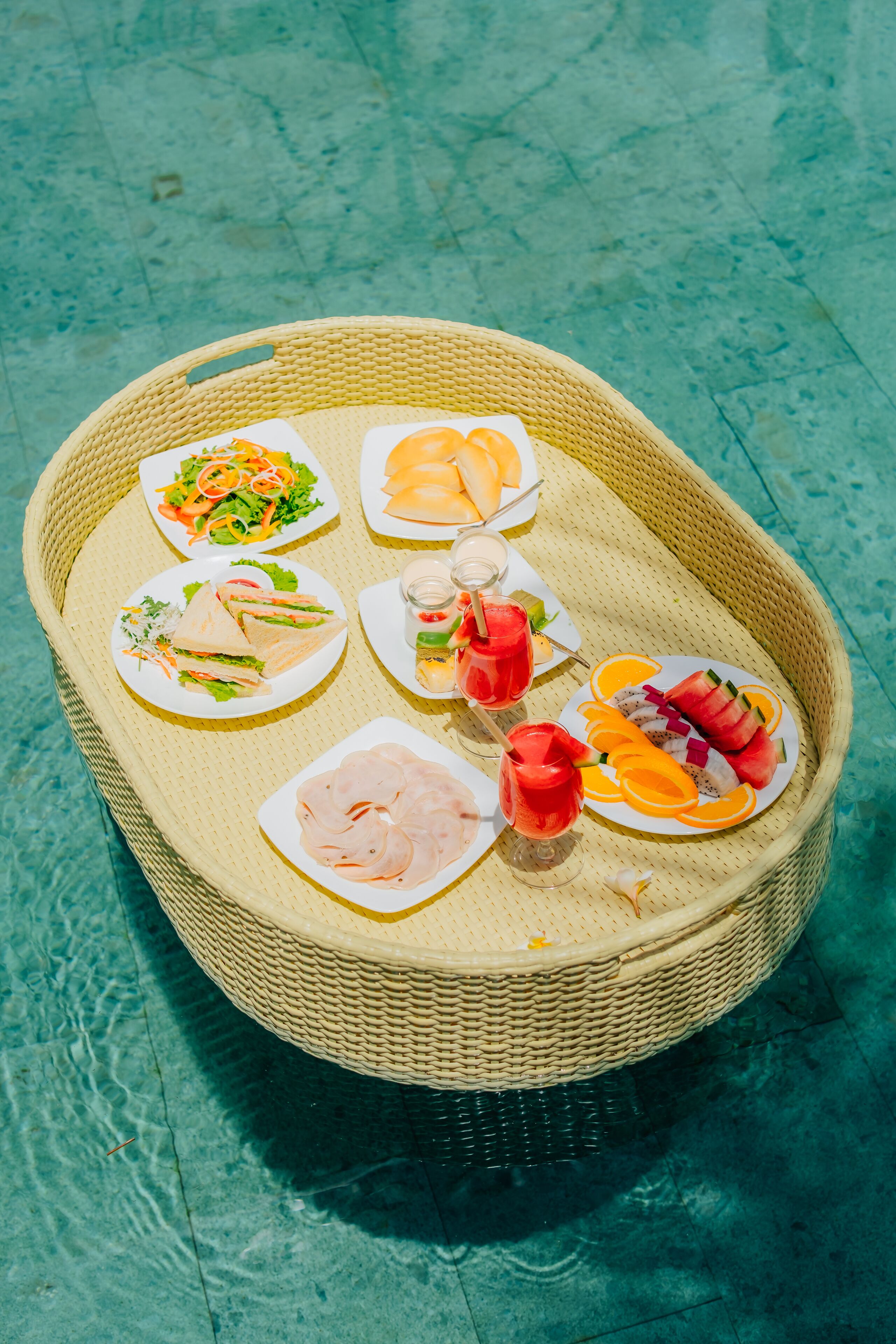 Uma bandeja de vime com diversos alimentos e bebidas sobre uma mesa junto à piscina.