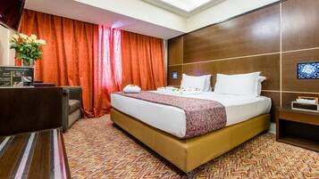 Quarto de hotel com cama, escrivaninha, cadeira, mesa de cabeceira e janela com cortinas.