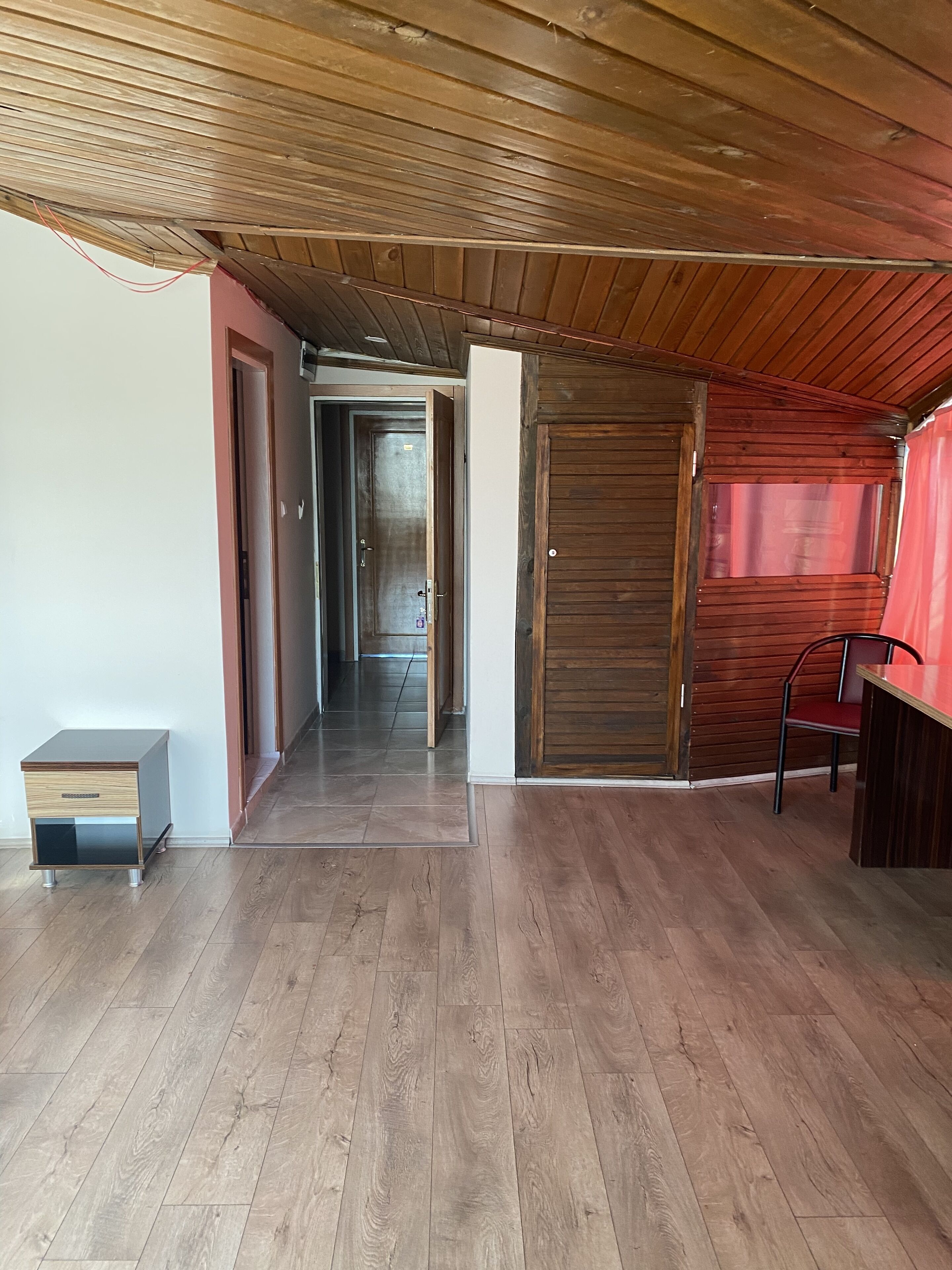 Quarto com piso de madeira, uma pequena mesa de cabeceira de madeira, uma cortina vermelha e uma porta que dá para outro cômodo.