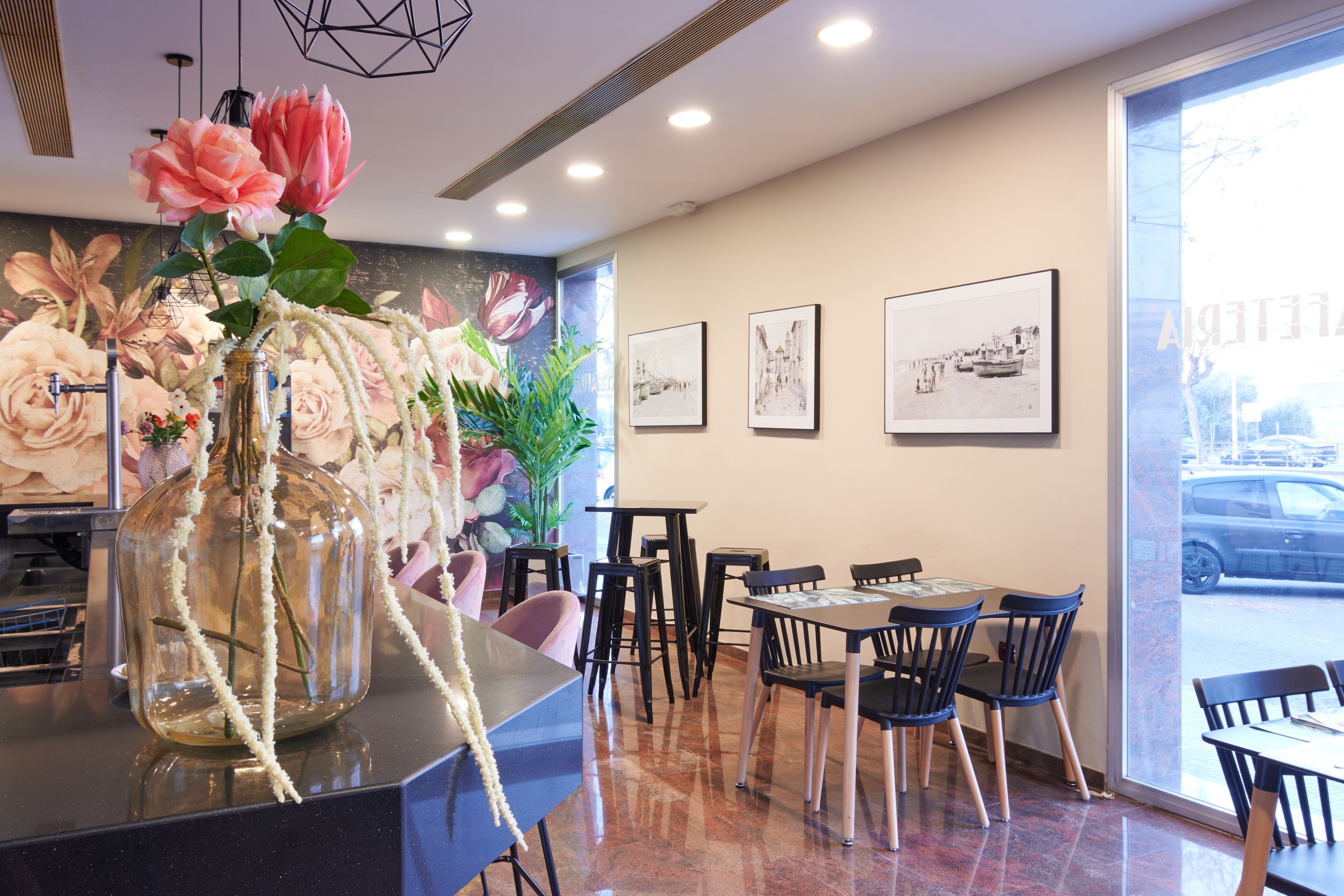 Interior de restaurante com balcão, cadeiras e mesas. Há quadros nas paredes e um vaso com flores no balcão.