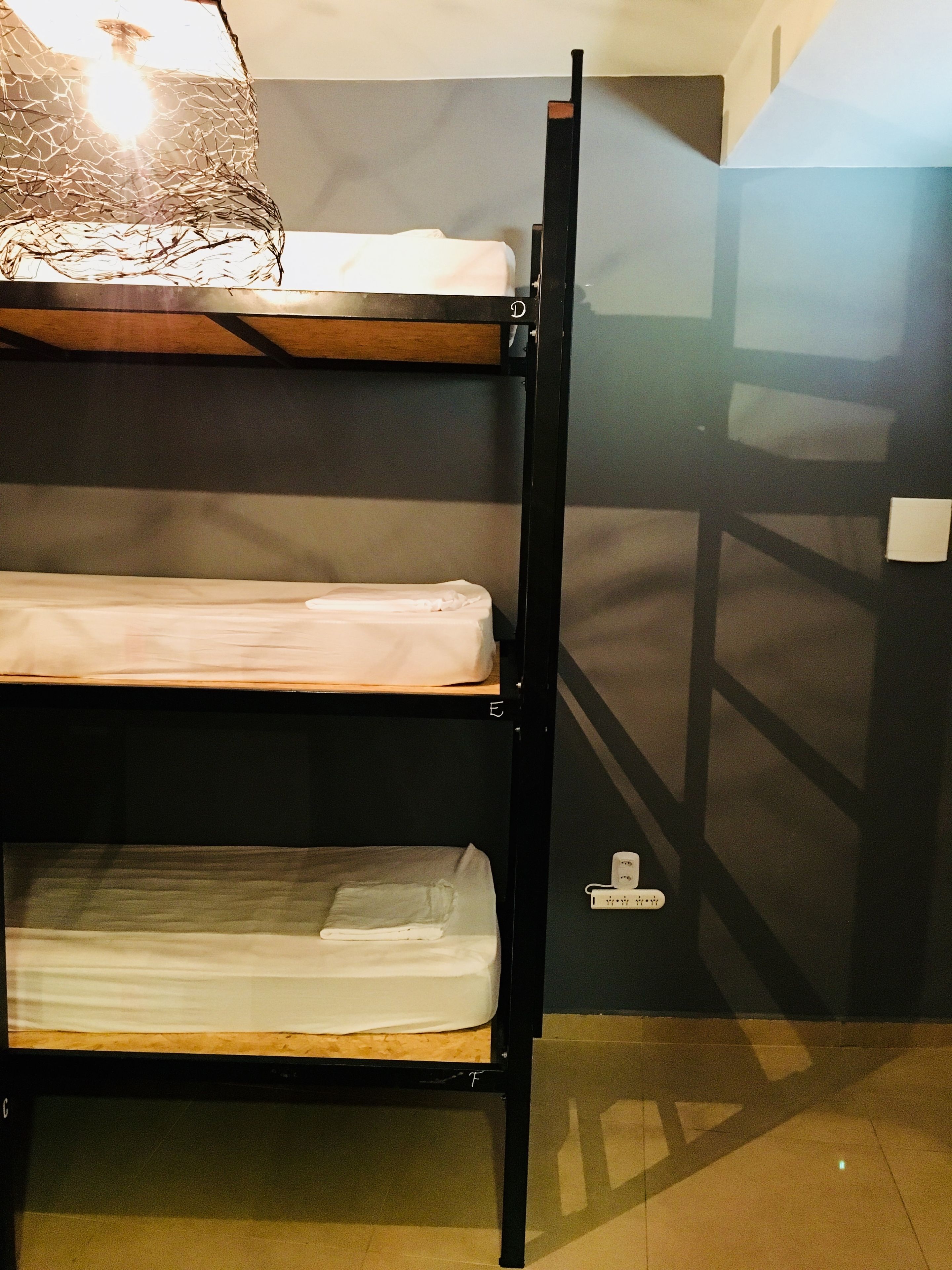 Uma cama beliche com três níveis, uma mesa pequena e um ponto de luz fixado na parede.