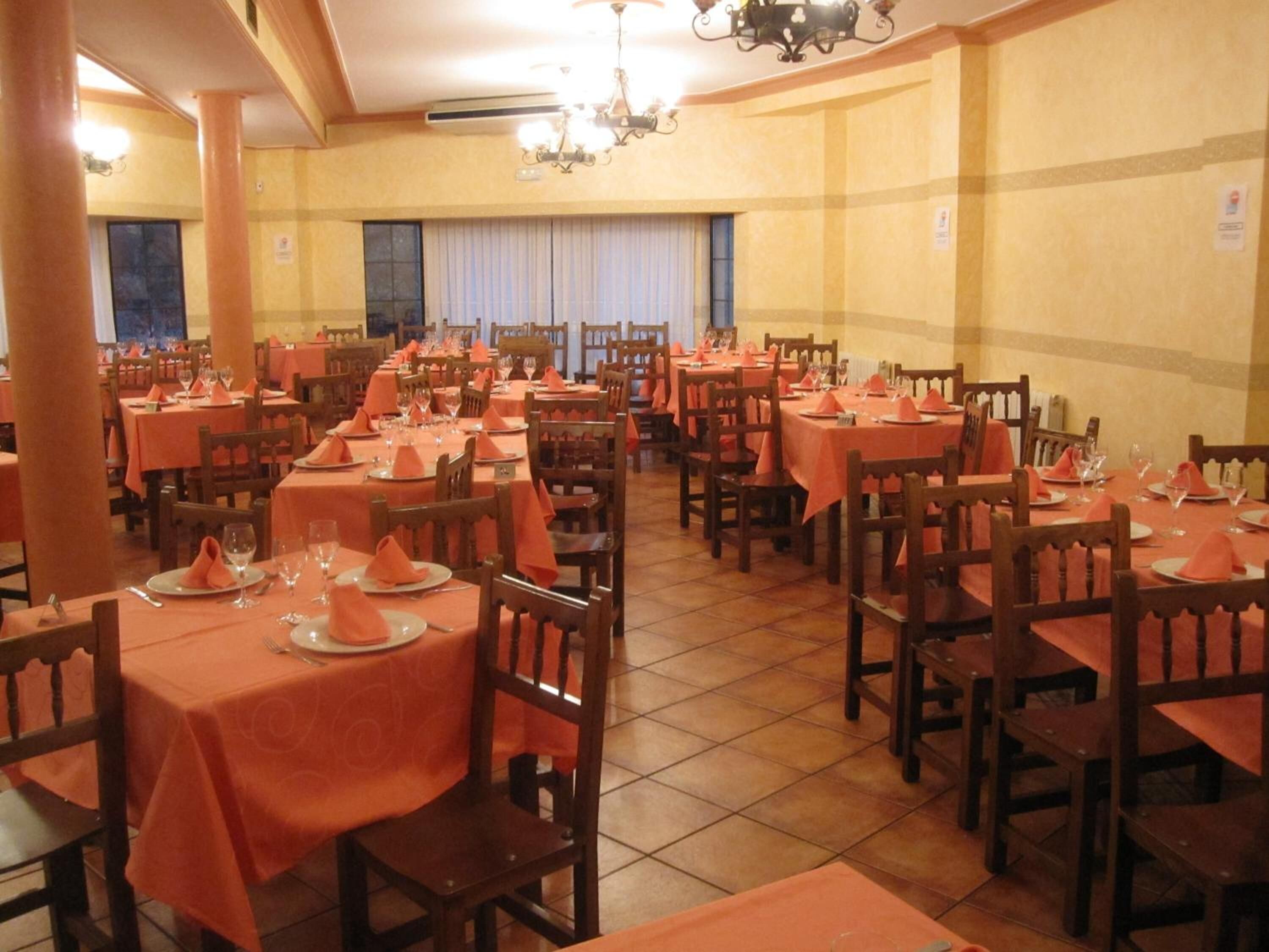 Uma sala de jantar com várias mesas dispostas, com toalhas e guardanapos laranja, cadeiras de madeira e um lustre.