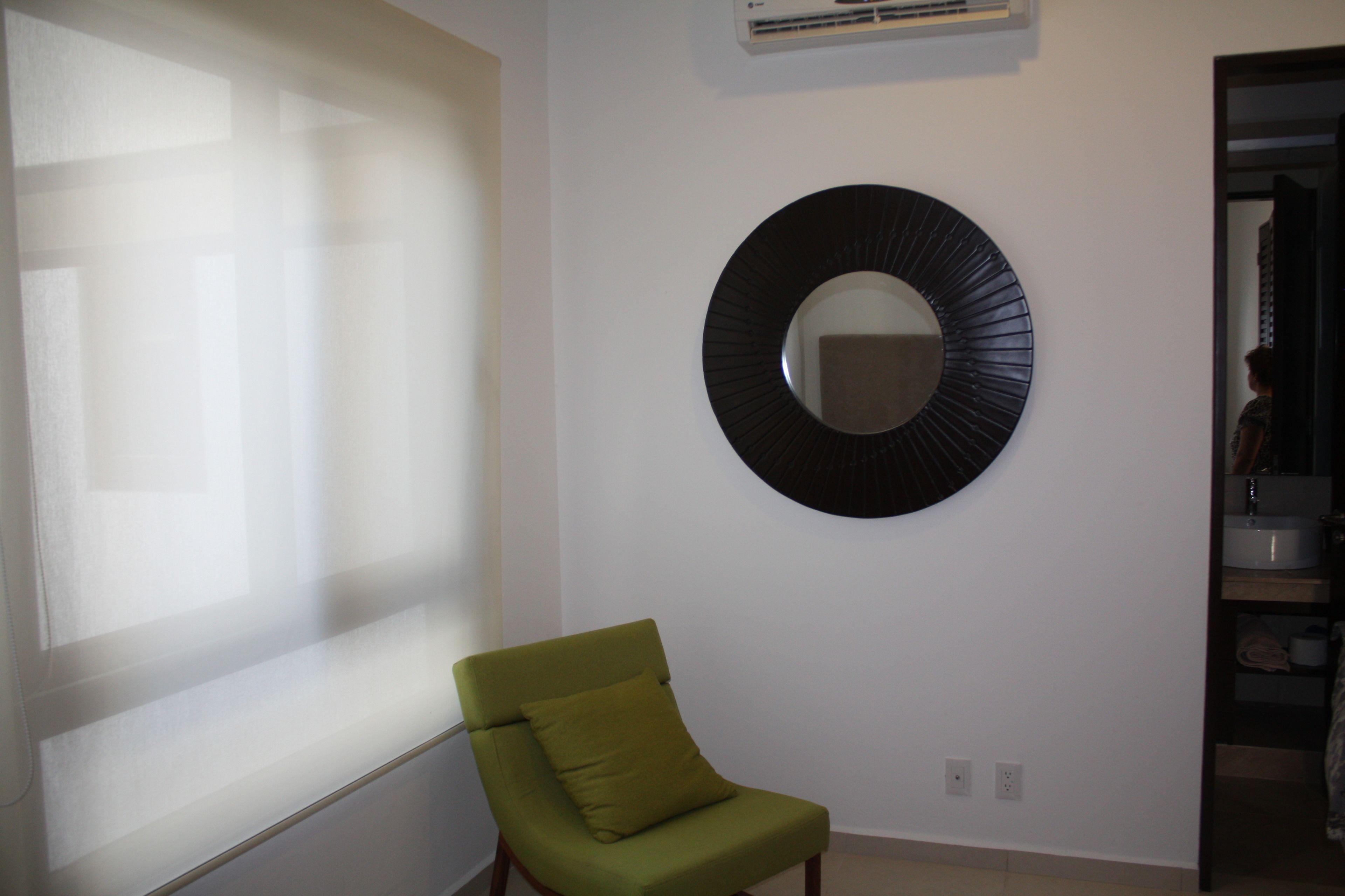 Quarto com uma cadeira verde, um espelho redondo na parede e uma janela com persianas.