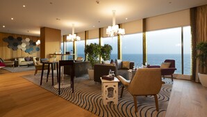Uma sala de estar espaçosa com uma grande janela com vista para o mar, equipada com sofás modernos, uma mesa de centro e plantas decorativas.