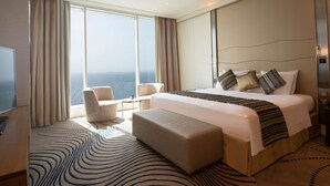 Quarto de hotel com uma cama grande, um sofá, uma cadeira e uma televisão. O quarto tem vista para o oceano.