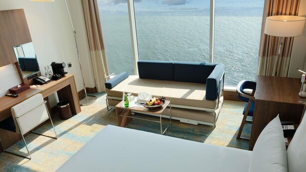 Quarto de hotel moderno com cama, escrivaninha, sofá e vista para o mar.