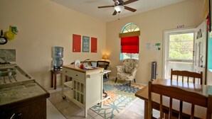 Cozinha com dispenser de água, ventilador de teto, espaço de jantar com mesa e cadeiras, e janela com cortinas vermelhas.