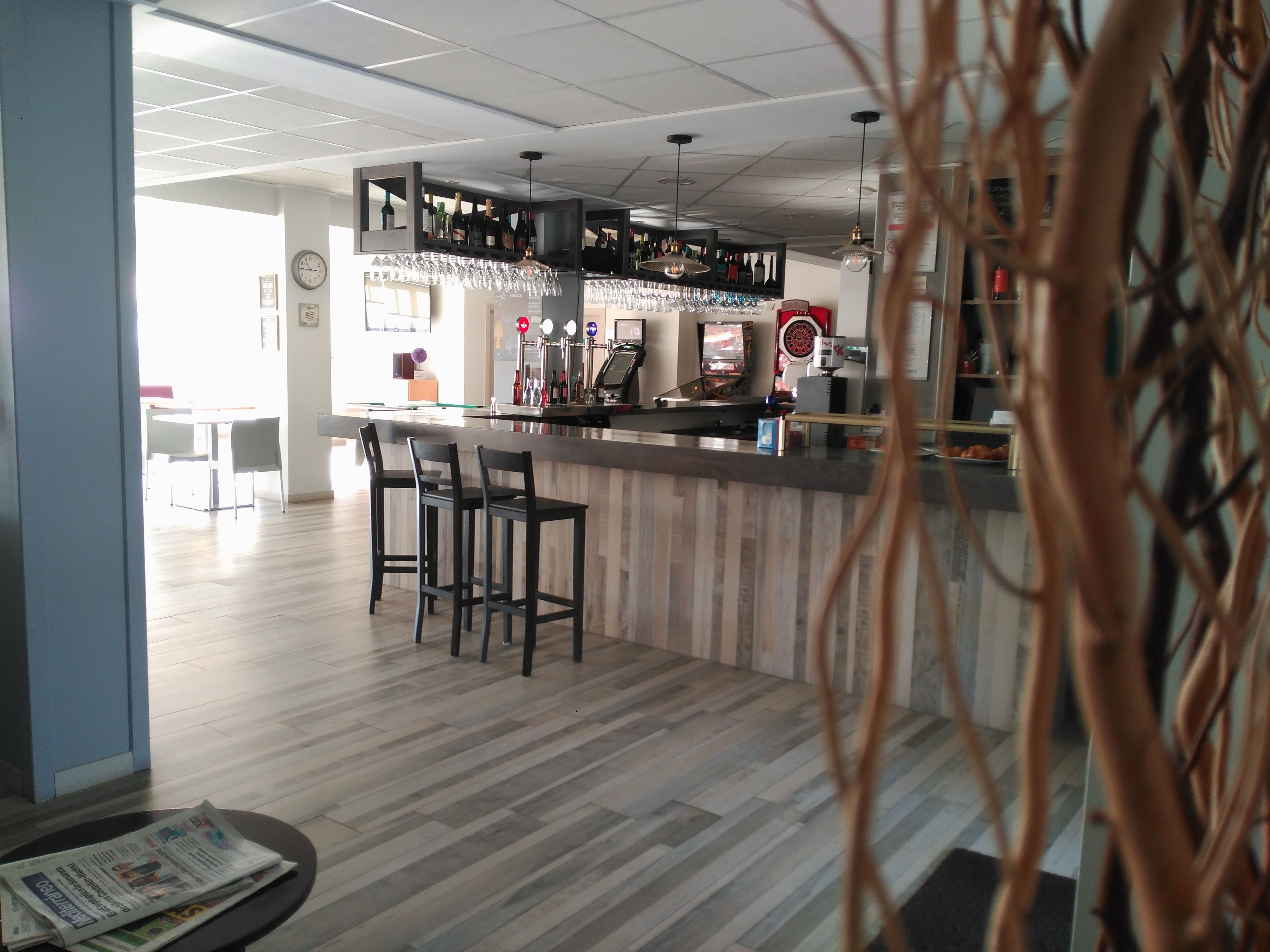 Uma área de bar com piso de madeira, um balcão com banquetas e diversas garrafas e copos em prateleiras.