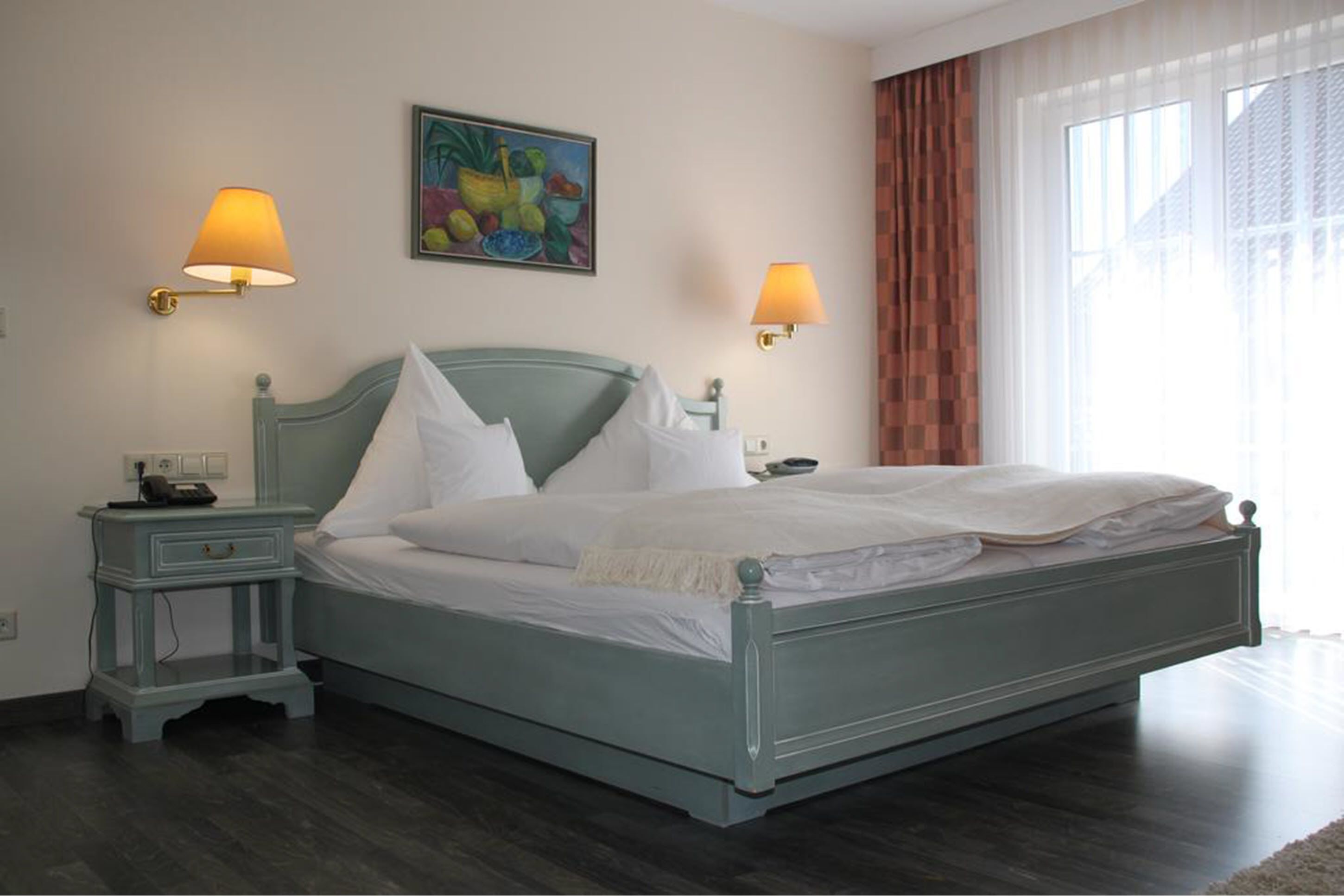 Cama bem arrumada com lençóis brancos, duas luminárias de cabeceira, mesa de cabeceira com telefone e um quadro na parede.