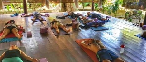 Um grupo de pessoas deitadas em tapetes de yoga em área externa coberta, com piso de madeira e luz natural entrando.