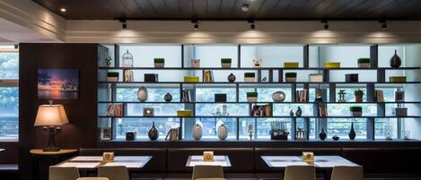 Um restaurante com várias mesas e cadeiras, uma estante com diversos objetos decorativos e vista para o exterior.