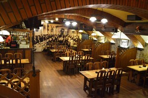 Um restaurante com mesas e cadeiras de madeira, área de bar com balcão e um mural na parede.