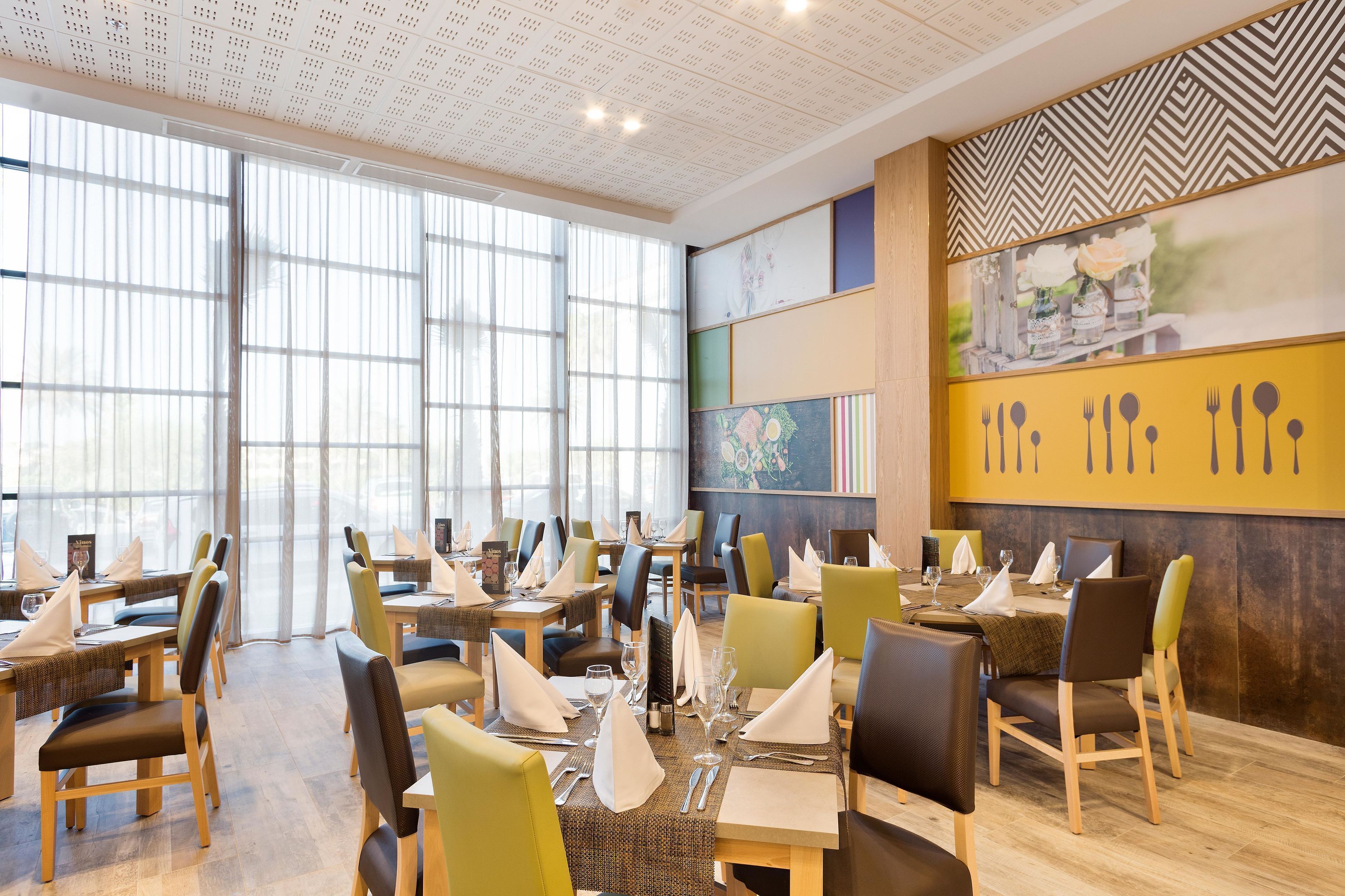 Um restaurante com várias mesas e cadeiras, grandes janelas e obras de arte nas paredes.