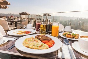 Mesa de café da manhã com pratos de comida, um copo de suco de laranja e acompanhamentos, com vista.