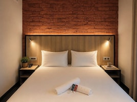 Quarto de hotel com parede de tijolos, cabeceira de madeira, duas almofadas brancas, cama bem arrumada, mesa de cabeceira com abajur, uma planta e uma toalha enrolada com artigos de higiene pessoal.