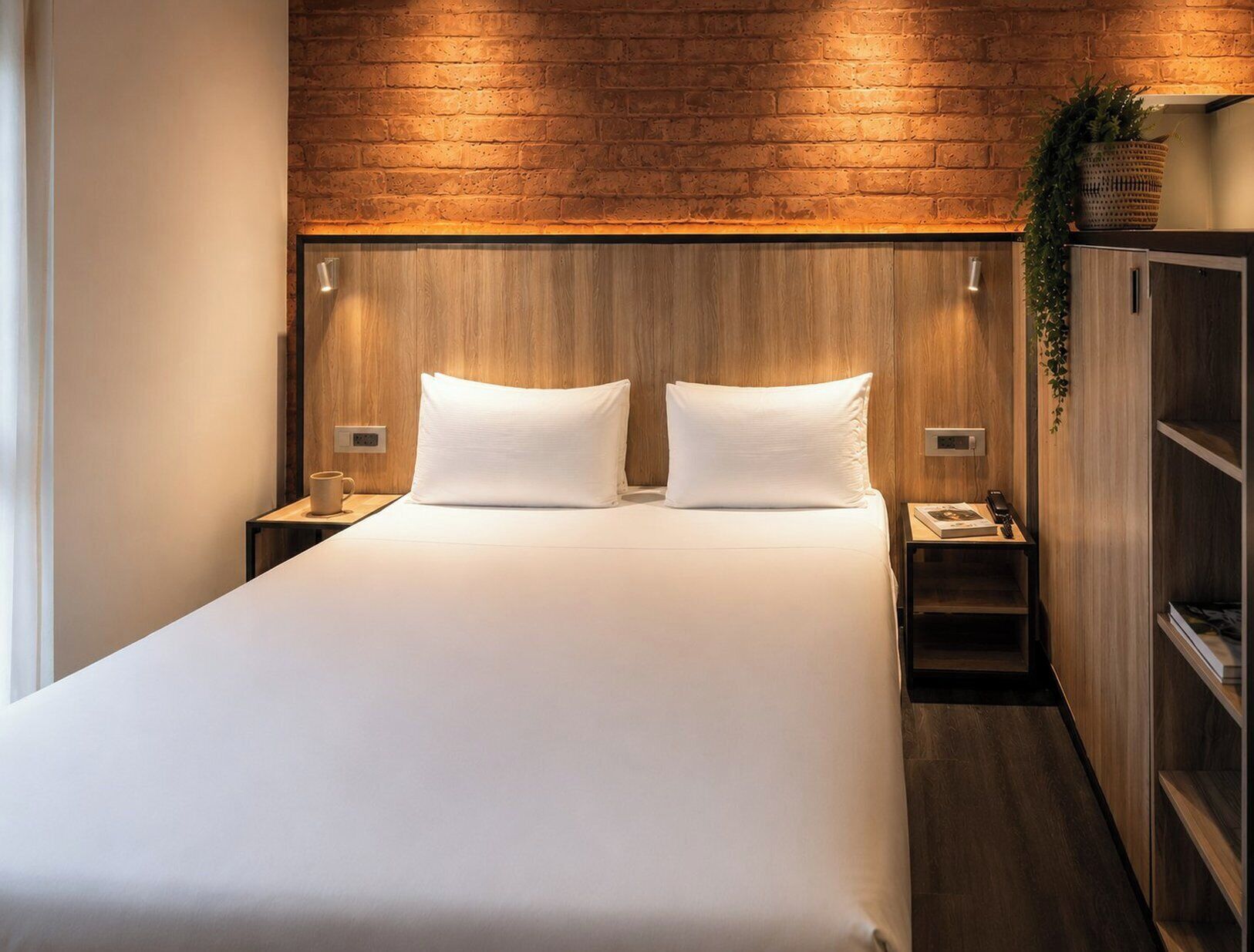 Quarto de hotel com cabeceira de madeira, uma cama grande com roupa de cama branca, mesas de cabeceira com abajures e uma planta no criado-mudo.