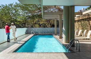 Piscina no terraço com superfície de água azul cristalina, rodeada por um deck de azulejos e uma área coberta com espreguiçadeiras.