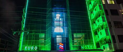 Um hotel com um letreiro chamativo 'EURO HOTEL', fachada de vidro e contorno de neon verde.