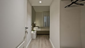 Um corredor estreito que dá para um quarto com cama, mesa de cabeceira e um cabideiro.