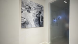 Um corredor com uma grande fotografia em preto e branco na parede, uma porta de vidro com a sinalização 'DOMUS ROMA' e um chão de cor clara.