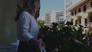 Uma mulher está perto de uma varanda, com vista para edifícios e um jardim.