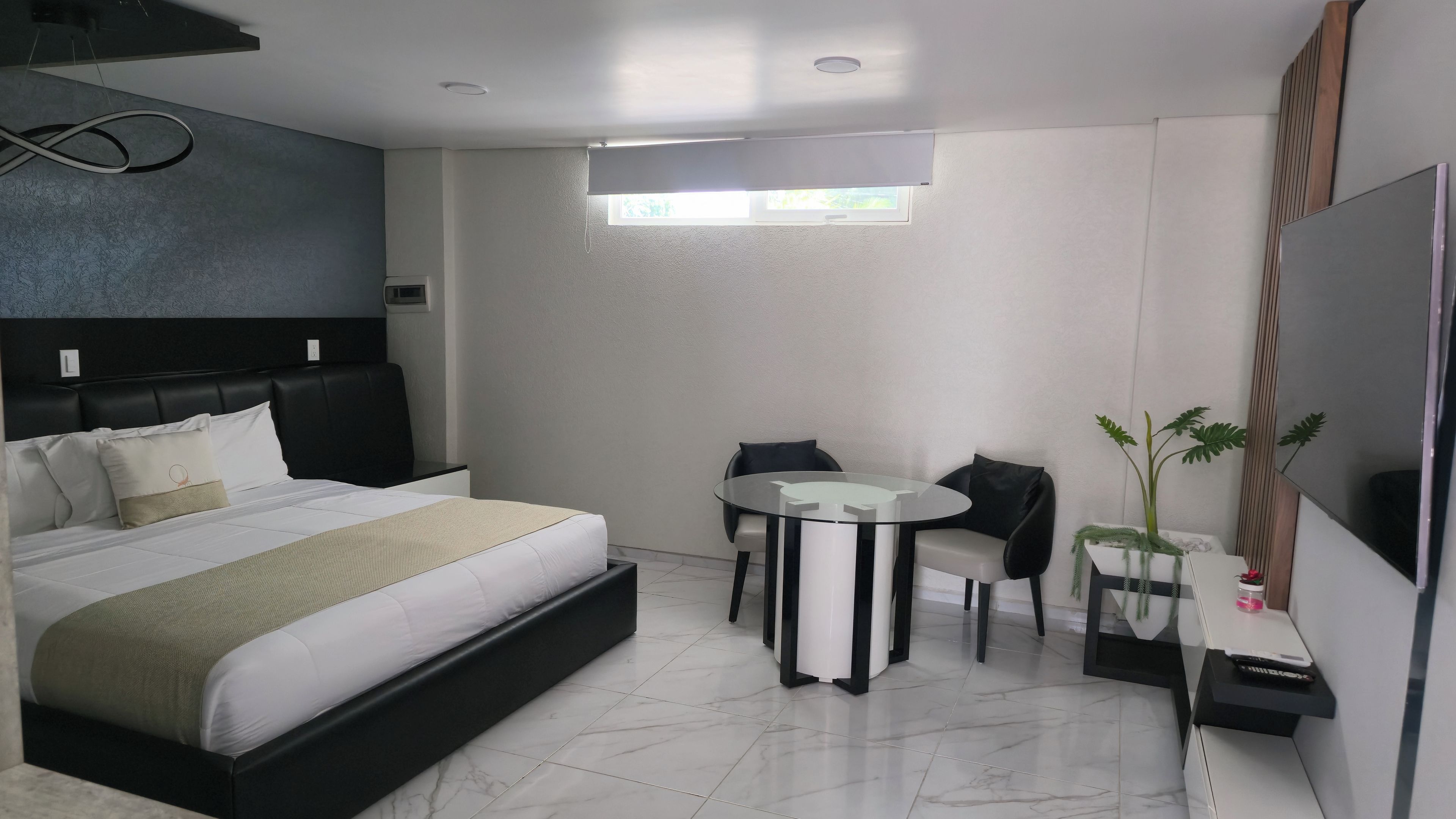 Quarto de hotel moderno com uma cama grande, uma mesa redonda, duas cadeiras, uma televisão e um vaso de planta.