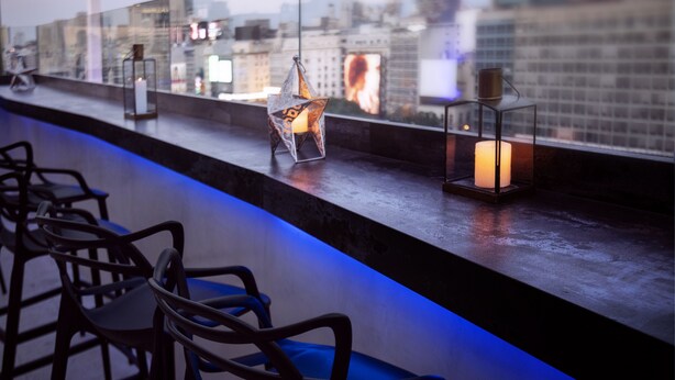 Uma área de bar com cadeiras e mesas, vista da cidade através de um parapeito de vidro e lanternas decorativas.