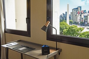 Mesa com luminária, papéis e uma caixa, com vista para um horizonte urbano e um monumento imponente.