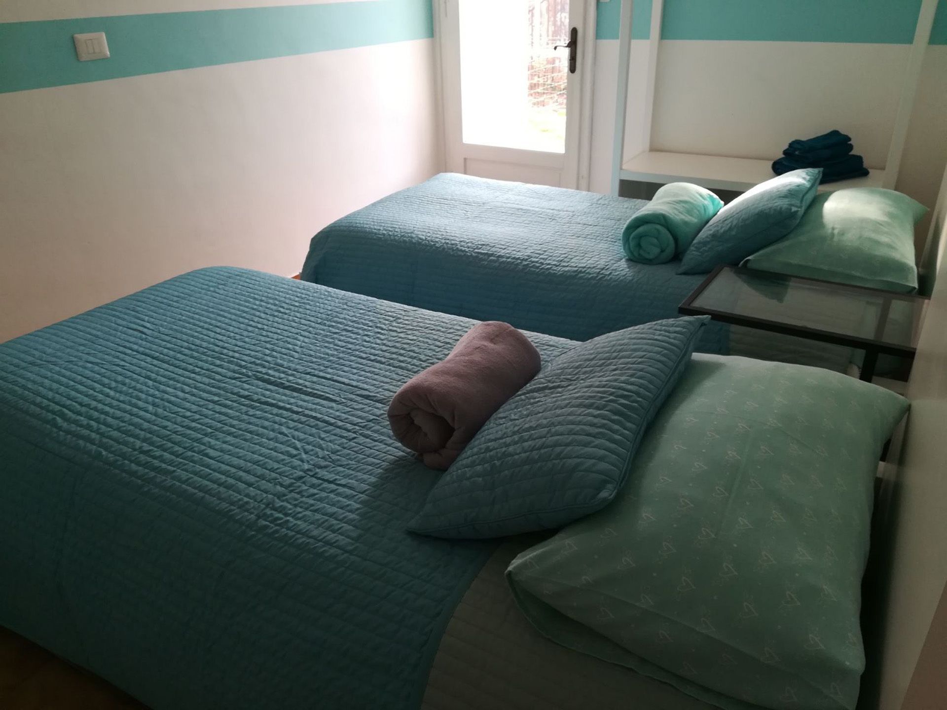 Quarto com duas camas, cada uma com colcha e almofadas azul-escuras. Há uma janela com vista para o exterior e uma porta à direita.