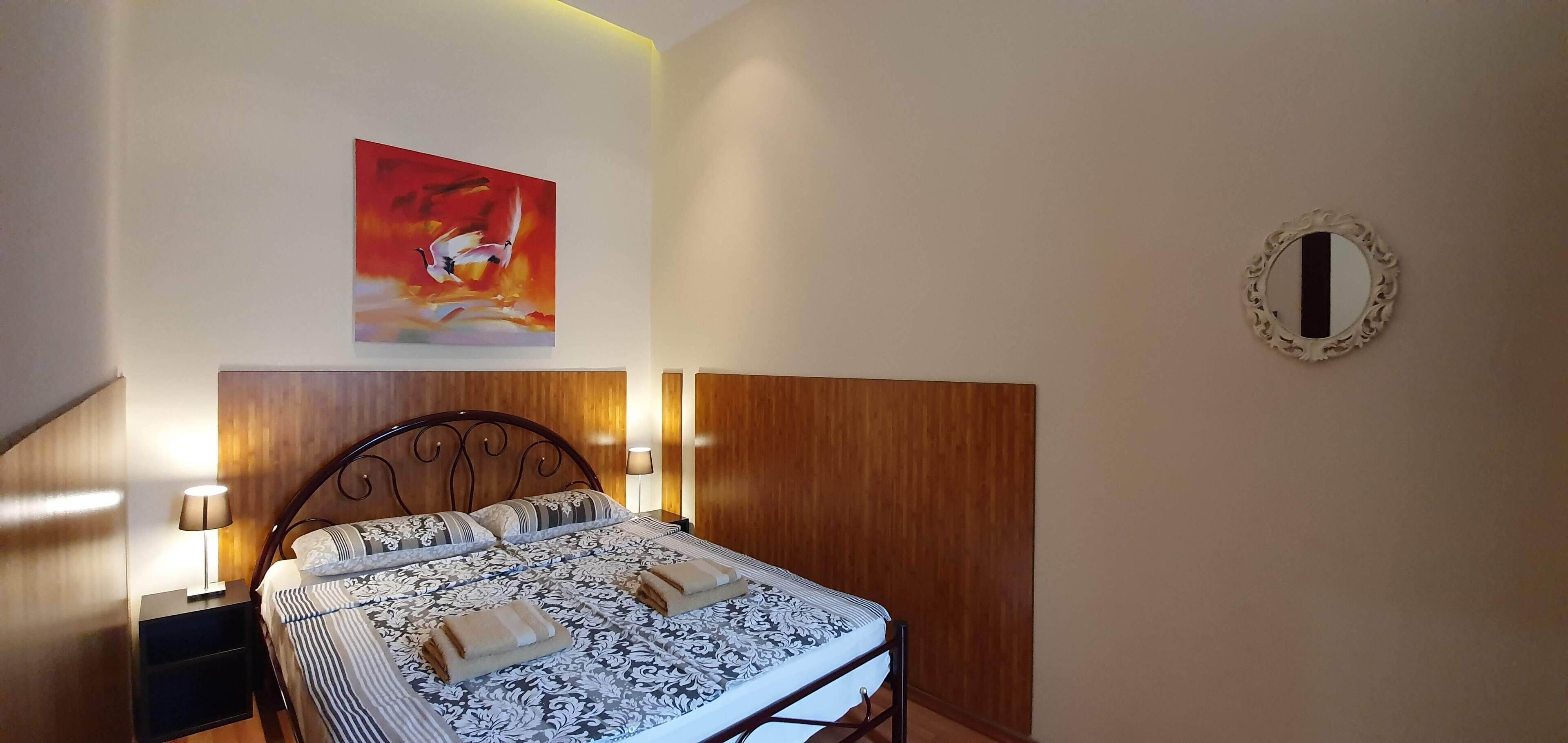 Uma cama bem arrumada com um edredom estampado e duas toalhas, um abajur, um espelho fixado na parede e uma pintura de um pássaro em voo.