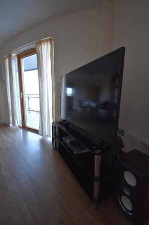 Sala de estar com uma grande TV de tela plana em um suporte, piso de madeira, uma porta de vidro deslizante que dá para uma área externa e um pequeno alto-falante preto à direita.