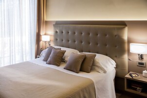 Quarto de hotel com uma cama grande, cabeceira, mesa de cabeceira com abajur e outra mesa com um livro e um recipiente.
