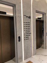 Um hall de hotel com piso e paredes de mármore, apresentando um elevador com visor digital e um diretório fixado na parede listando quartos de hóspedes, restaurante, centro de fitness, área de lobby/recepção e estacionamento.