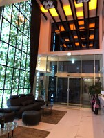 Um lobby moderno com ampla entrada de porta de vidro, área de estar com sofás e mesa de centro, e teto com vigas aparentes e iluminação embutida.