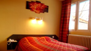 Quarto de hotel com cama, telefone de apoio, luminária na parede, radiador e janela com cortinas.