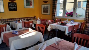 Restaurante com várias mesas, cada uma coberta por um pano de mesa branco e cadeiras de madeira. Há quadros nas paredes e uma janela com vista para o exterior.