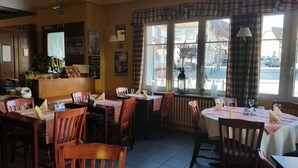 Interior de restaurante com mesas e cadeiras de madeira, um balcão com menu e vista para a rua através de grandes janelas.