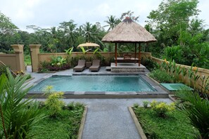 Piscina com espreguiçadeiras, um gazebo com telhado de palha e vegetação tropical exuberante.