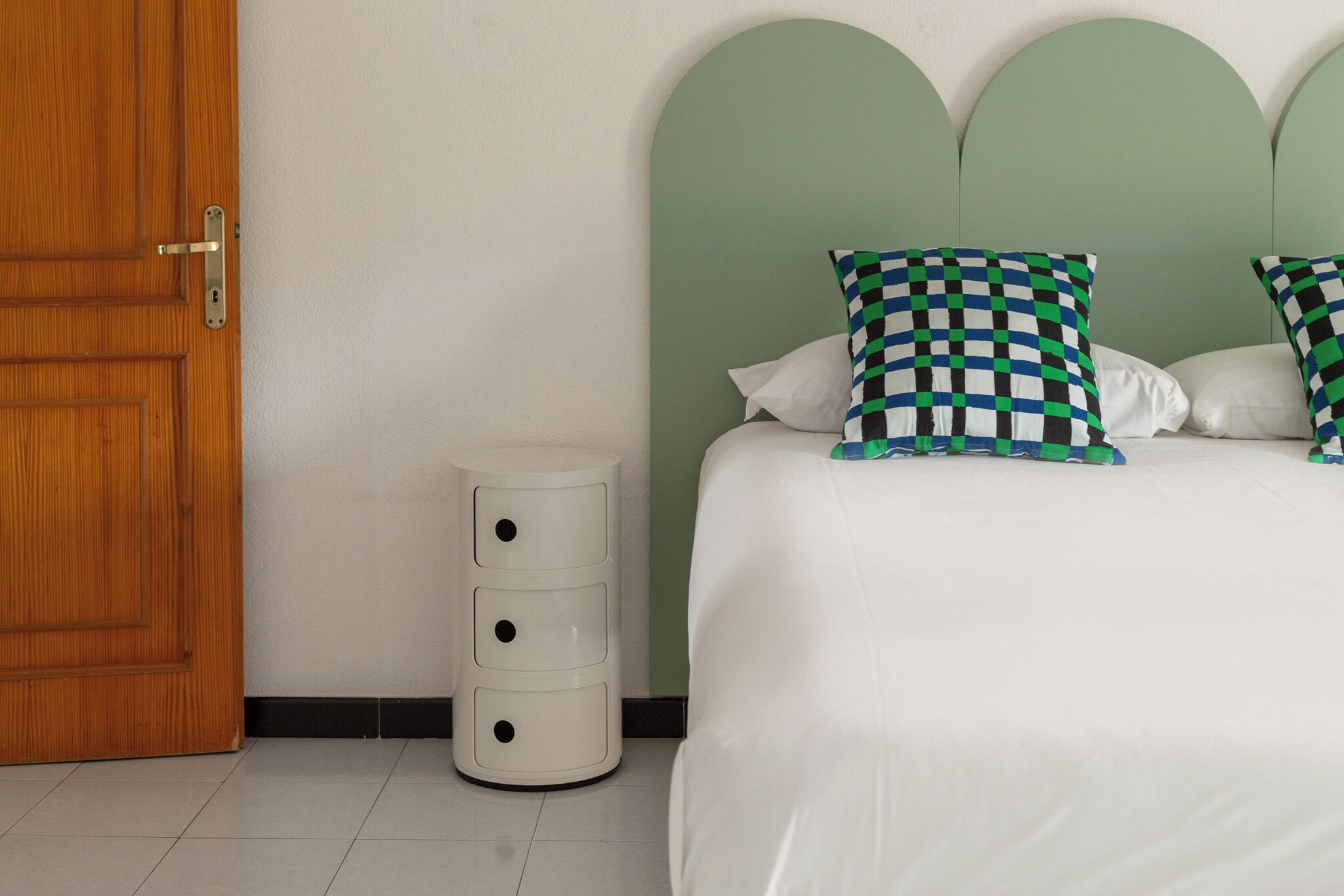 Quarto com porta de madeira, mesa de cabeceira branca com gavetas e cama com almofadas de padrão xadrez verde e branco.