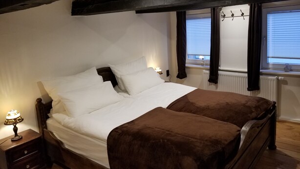 Quarto com cama de madeira, roupa de cama branca, mesa de cabeceira com abajur e janela com cortinas.