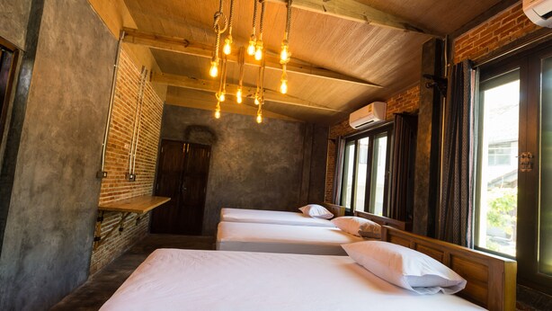 Quarto de hotel com três camas, vigas de madeira, paredes de tijolo e luminárias pendentes.
