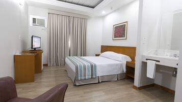 Quarto de hotel com cama, escrivaninha com computador, cadeira, banheiro com espelho e pia, e um quadro na parede.